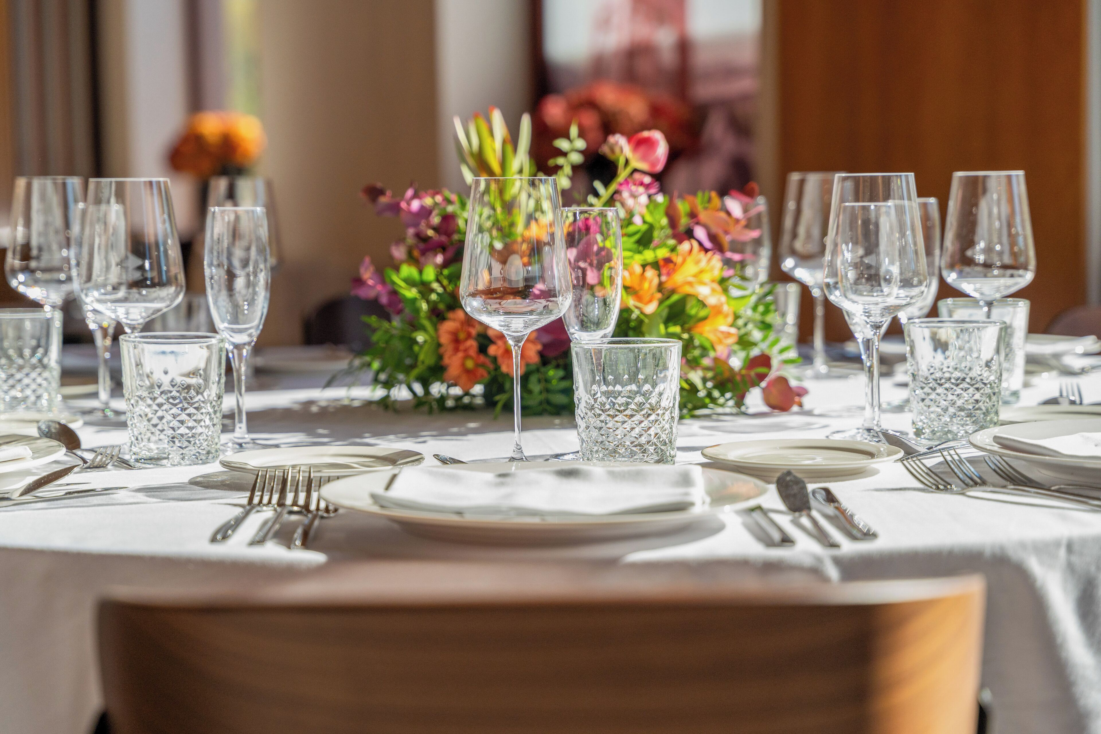 Uma mesa posta para uma experiência gastronômica requintada, com copos, pratos e talheres cristalinos, adornada por um arranjo floral.