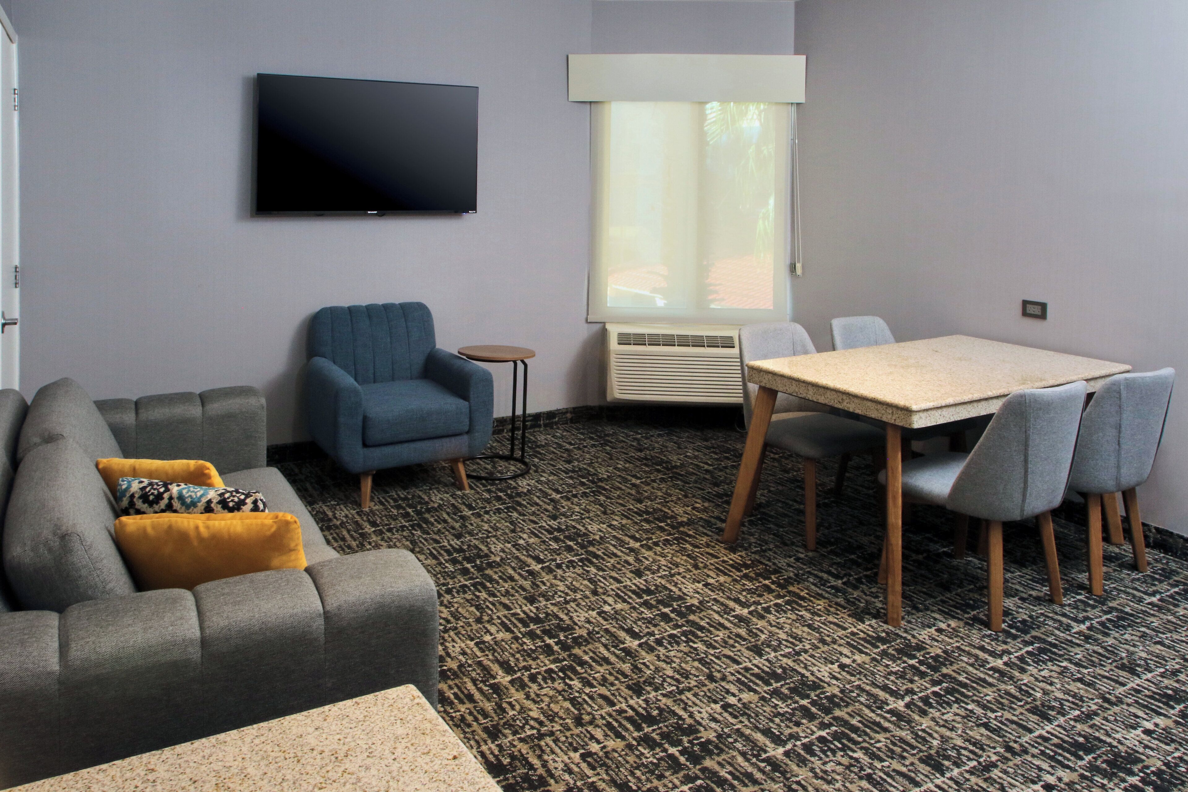 Quarto de hotel moderno com sofá cinza, poltrona azul, mesa redonda pequena, televisão fixada na parede e uma janela grande com persianas.
