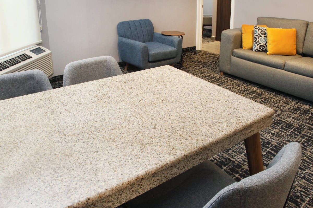 Quarto de hotel com mesa de granito, poltronas cinzas, sofá com almofadas amarelas e estampadas, e chão coberto por carpete.