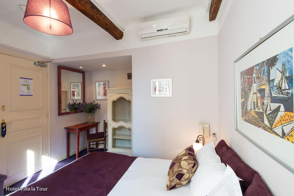 Quarto de hotel com cama, escrivaninha, cadeira, espelho, pintura e aparelho de ar condicionado.