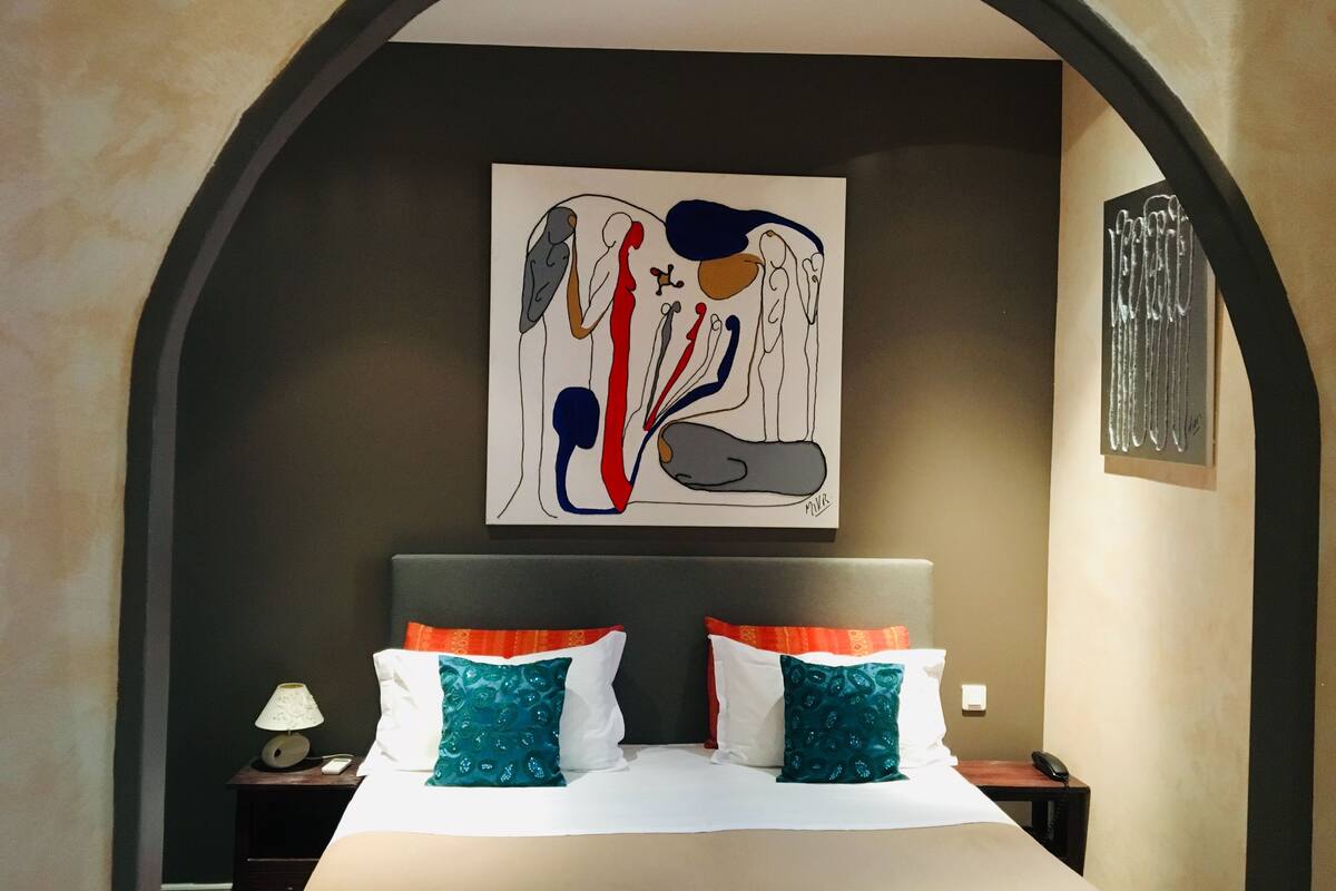 Quarto de hotel com uma cama grande, duas almofadas, uma mesa de cabeceira com abajur e um quadro na parede.