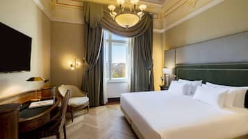 Quarto de hotel com uma cama grande, uma escrivaninha com cadeira, televisão de tela plana, janela com cortinas e um lustre.