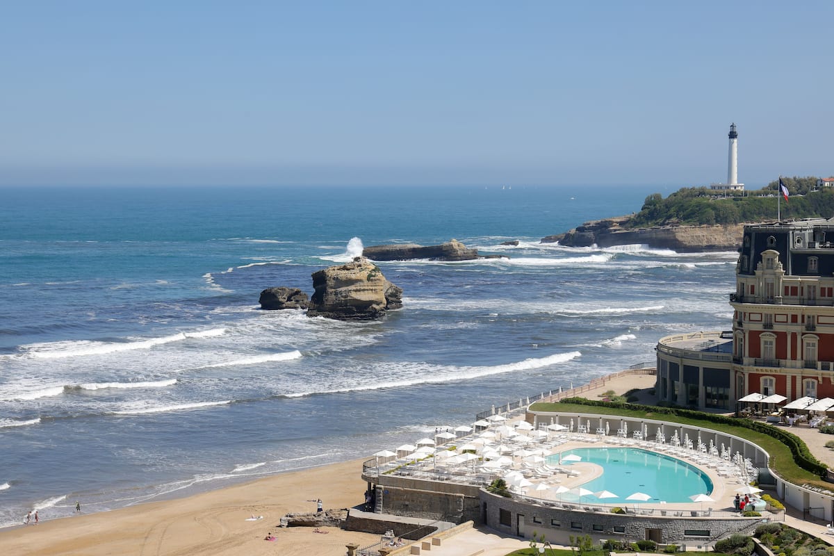 Um hotel à beira-mar com piscina, vista para o oceano e um farol ao fundo.
