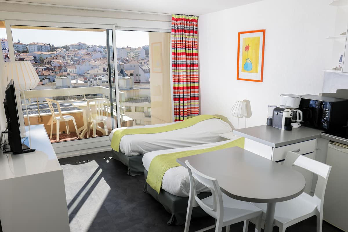 Quarto de hotel com cama, escrivaninha, cadeira, cafeteira e vista da cidade através de uma grande janela.