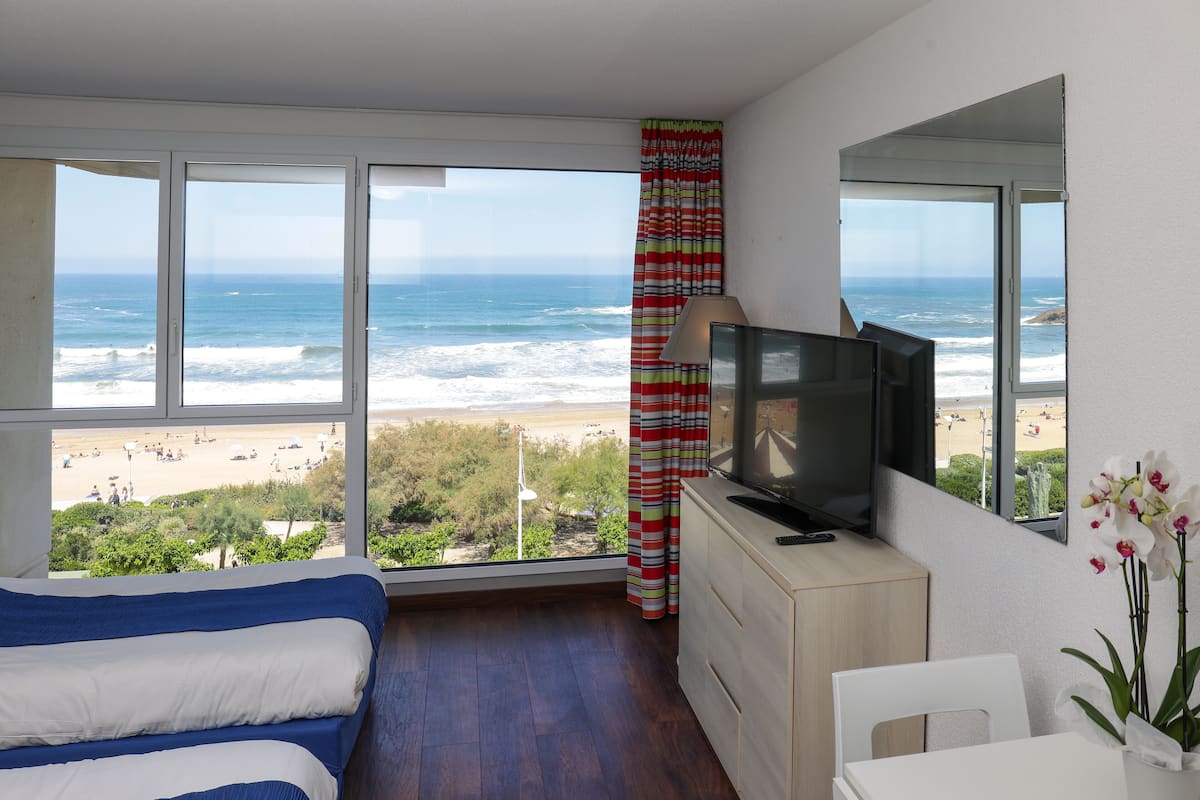 Quarto com uma grande janela com vista para a praia e oceano, uma cama, uma televisão e uma cômoda.