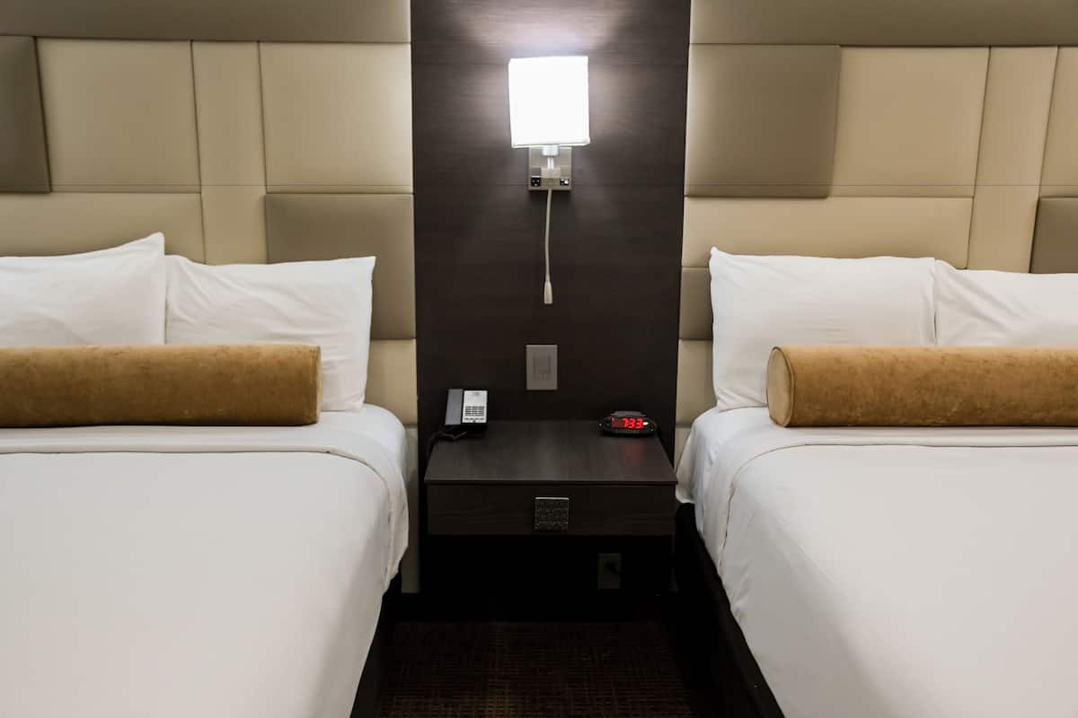 Quarto de hotel com duas camas, uma mesinha de cabeceira com telefone e relógio, e uma luminária na parede.