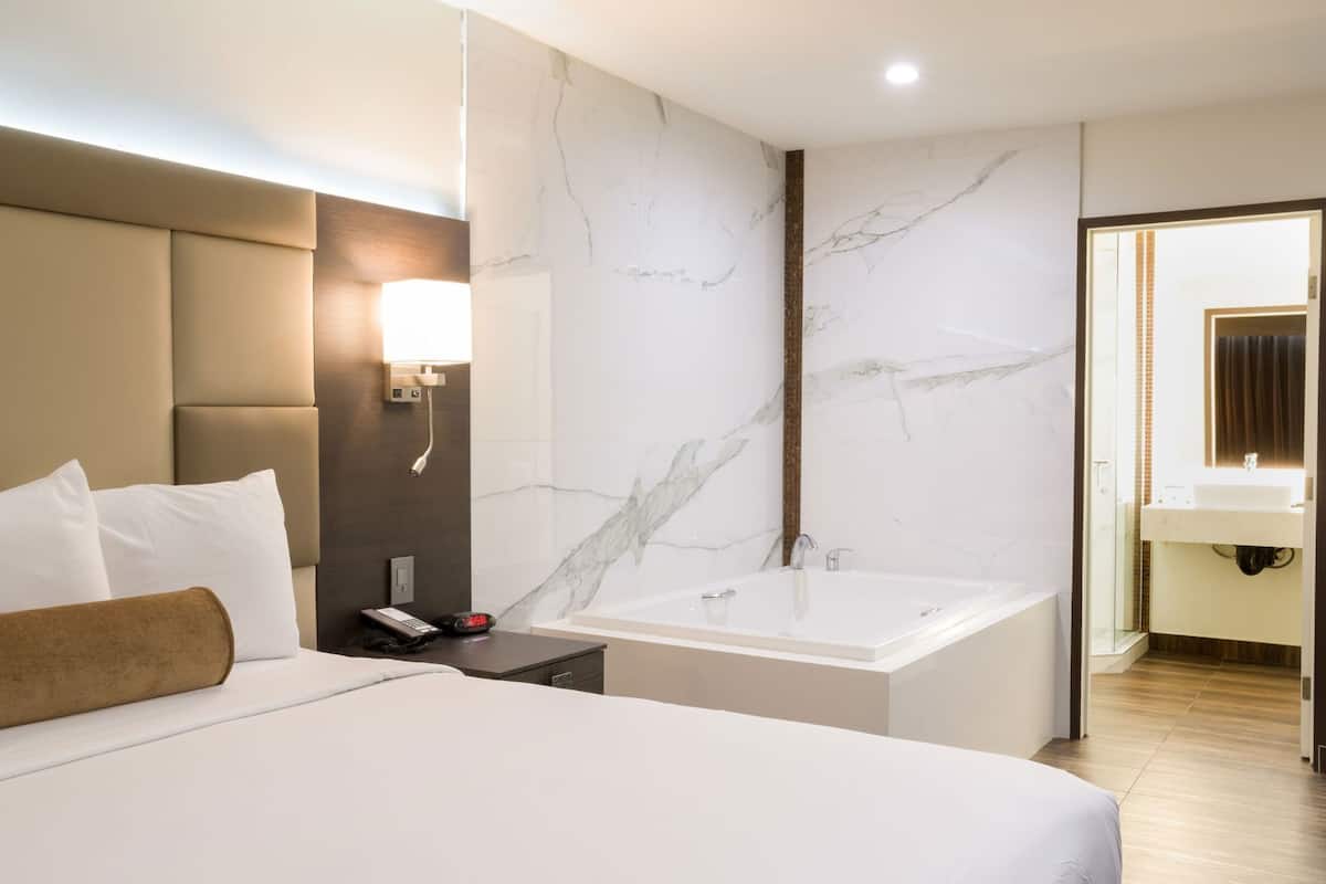 Quarto de hotel com uma cama grande, mesa de cabeceira com telefone e despertador, luminária fixada na parede, parede de mármore, banheira e banheiro com pia e espelho.