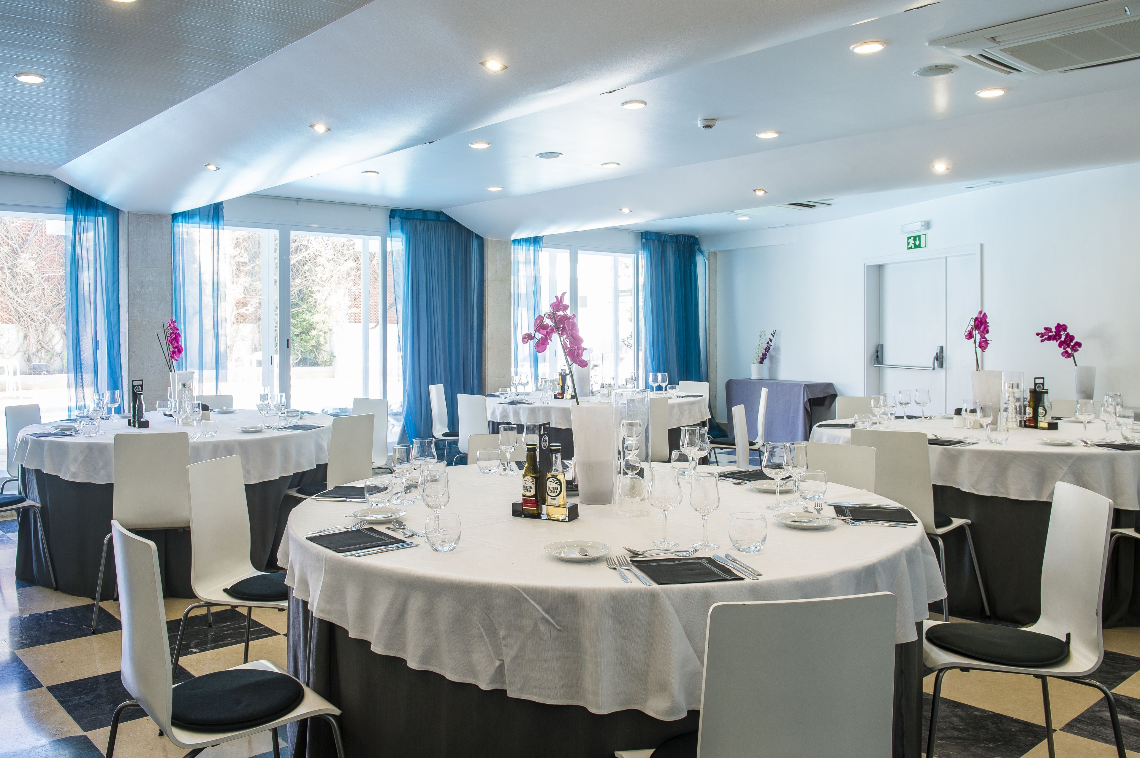 Sala de conferências com mesas redondas preparadas para refeições, equipada com cadeiras, pratos, talheres e copos de vinho.