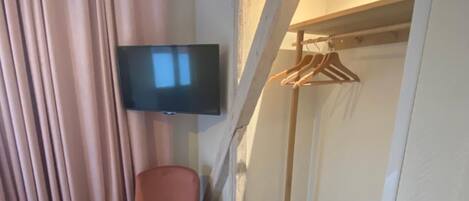 Quarto de hotel com televisão de tela plana na parede, uma cadeira, um porta-roupas e uma janela com cortinas.