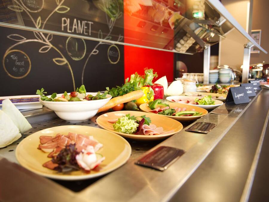 Buffet com diversos pratos, incluindo saladas e carnes, e uma lousa com a palavra 'PLANET' escrita.