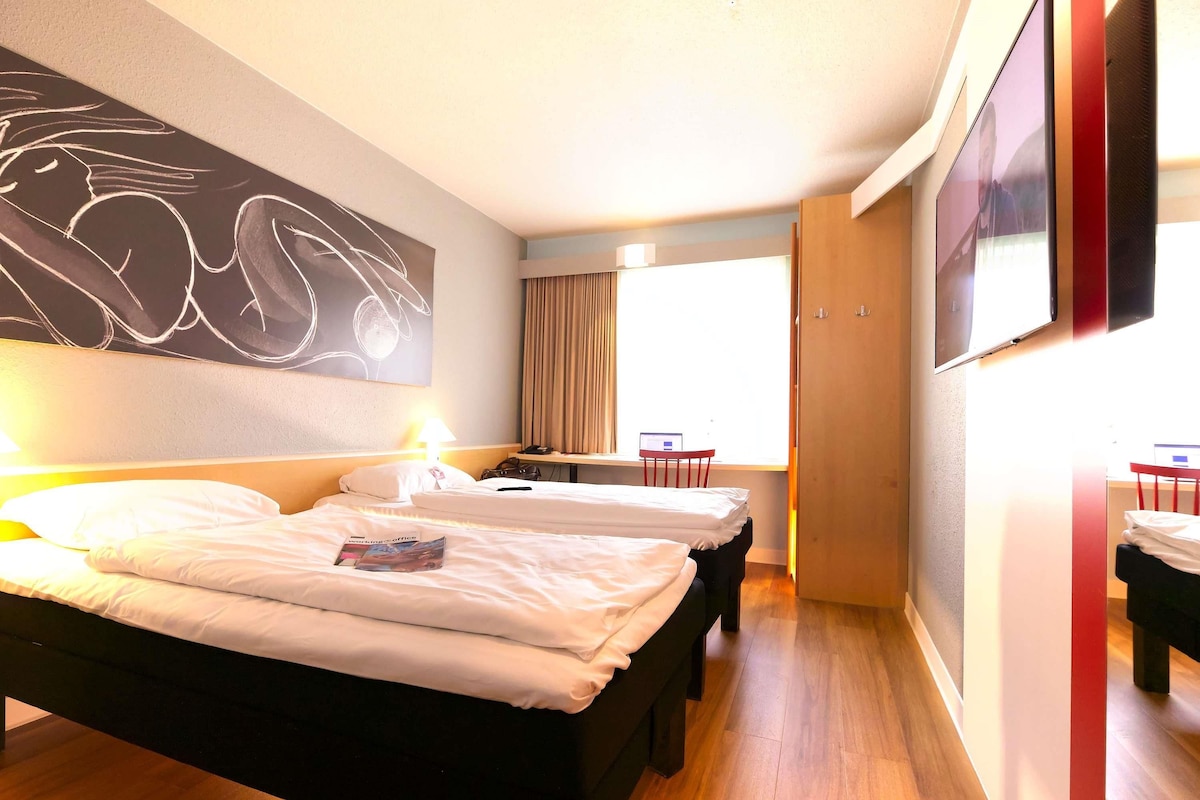 Quarto de hotel com duas camas, uma escrivaninha, uma cadeira, uma televisão e um quadro abstrato grande na parede.