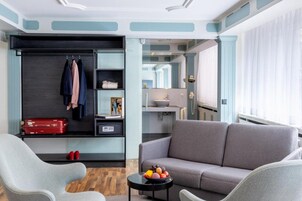 Quarto de hotel moderno com sofá cinza, uma mesa redonda pequena com frutas, um guarda-roupa com roupas penduradas e uma mala vermelha.