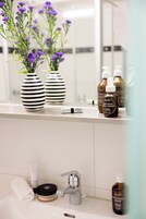 Um banheiro com espelho, dois vasos listrados com flores roxas e diversos produtos de cuidados naturais na bancada.