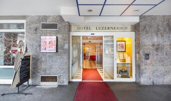 Entrada do Hotel Luzernhof com fachada de mármore, tapete vermelho e uma exposição de bolsas.