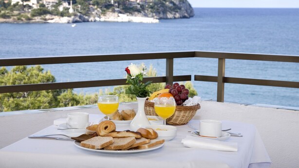 Mesa de café da manhã com croissants, pão e frutas, com vista para a costa e uma ilha ao longe.