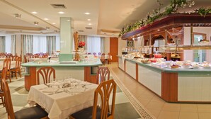 Restaurante com várias mesas e cadeiras, balcão de buffet com diversos alimentos e teto decorado com plantas penduradas.