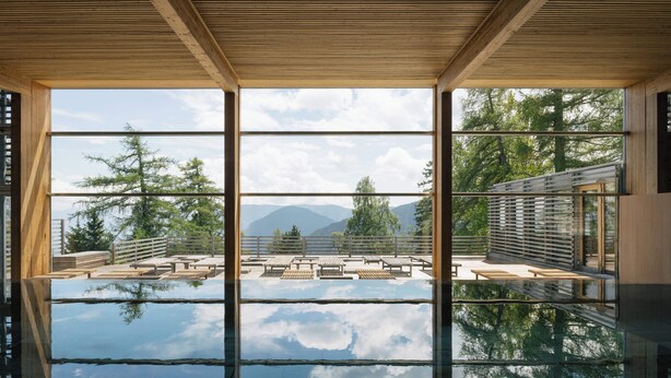 Uma área de piscina moderna com paredes de vidro, vigas de madeira e vista para montanhas e árvores.