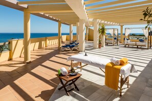 Área de spa ao ar livre banhada pelo sol, com mesa de massagem, toalhas e vista para o mar.