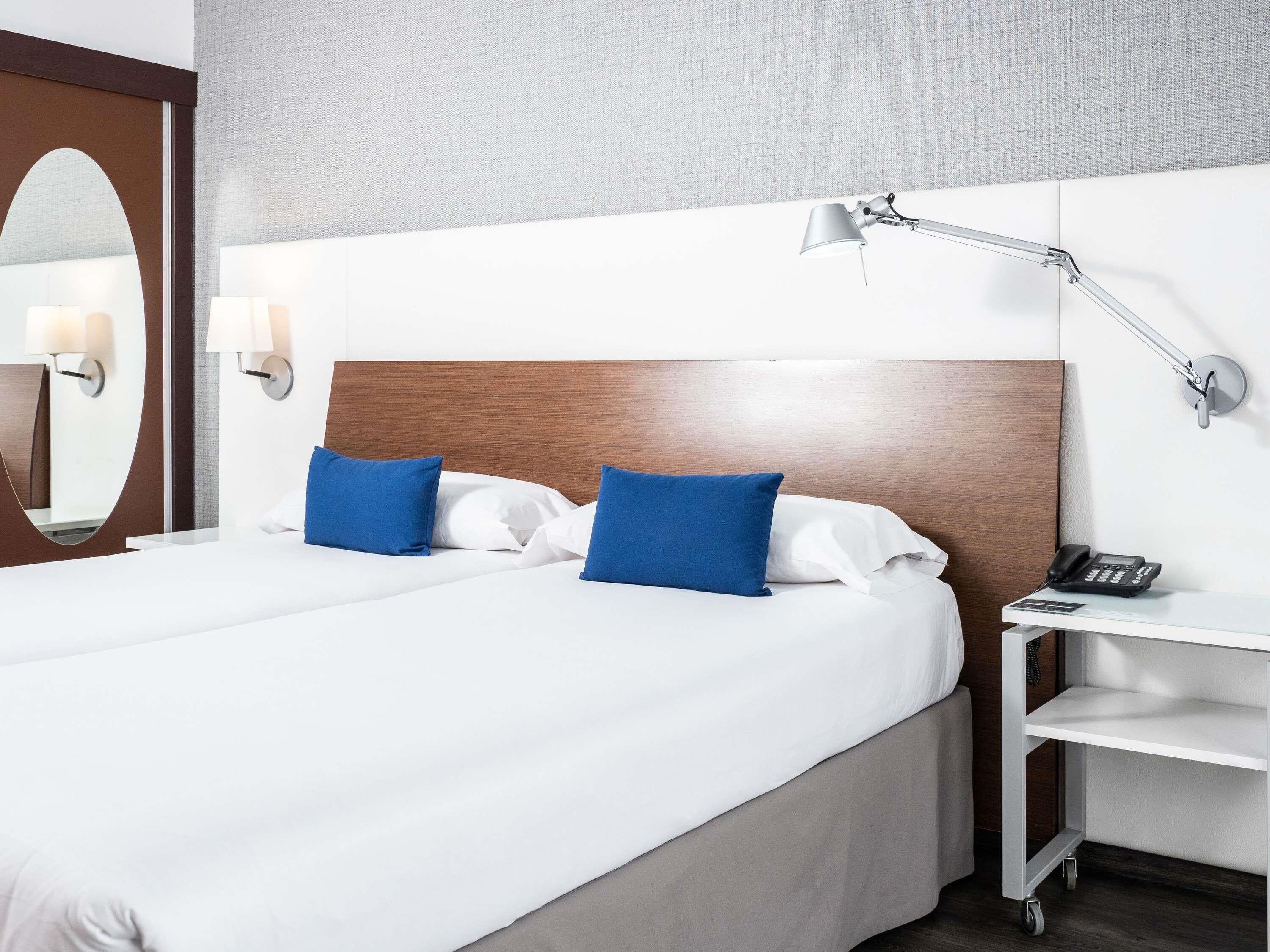 Quarto de hotel com uma cama grande, dois travesseiros azuis, cabeceira de madeira, mesa de cabeceira com telefone e luminária fixada na parede.
