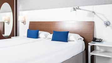 Quarto de hotel com uma cama grande, dois travesseiros azuis, cabeceira de madeira, mesa de cabeceira com telefone e luminária fixada na parede.