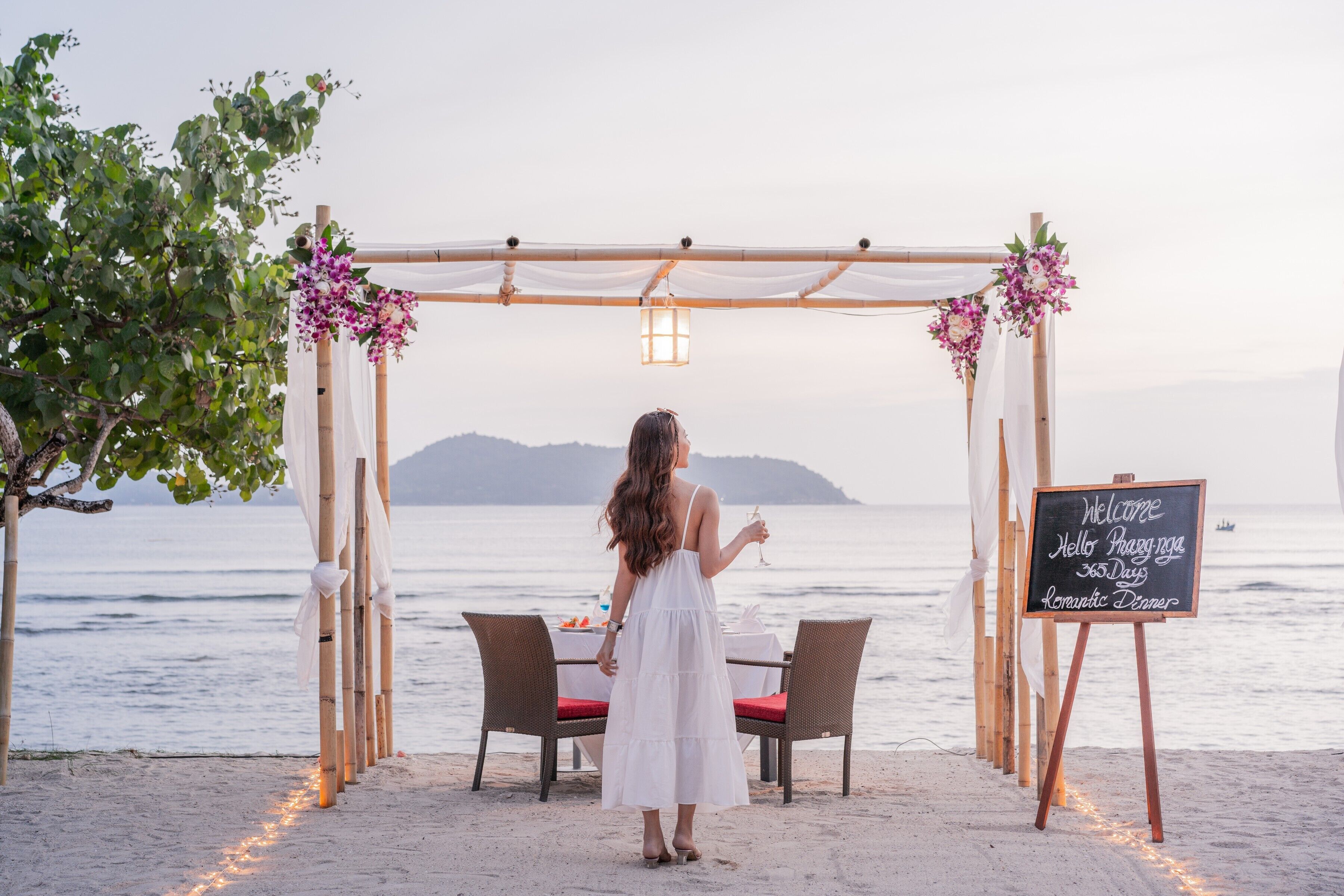 Montagem de refeições à beira-mar com um pérgola de madeira, cortinas brancas e decorações florais. Uma placa de lousa diz: "Welcome, Hello, 365 Days, Romantic Dinner".