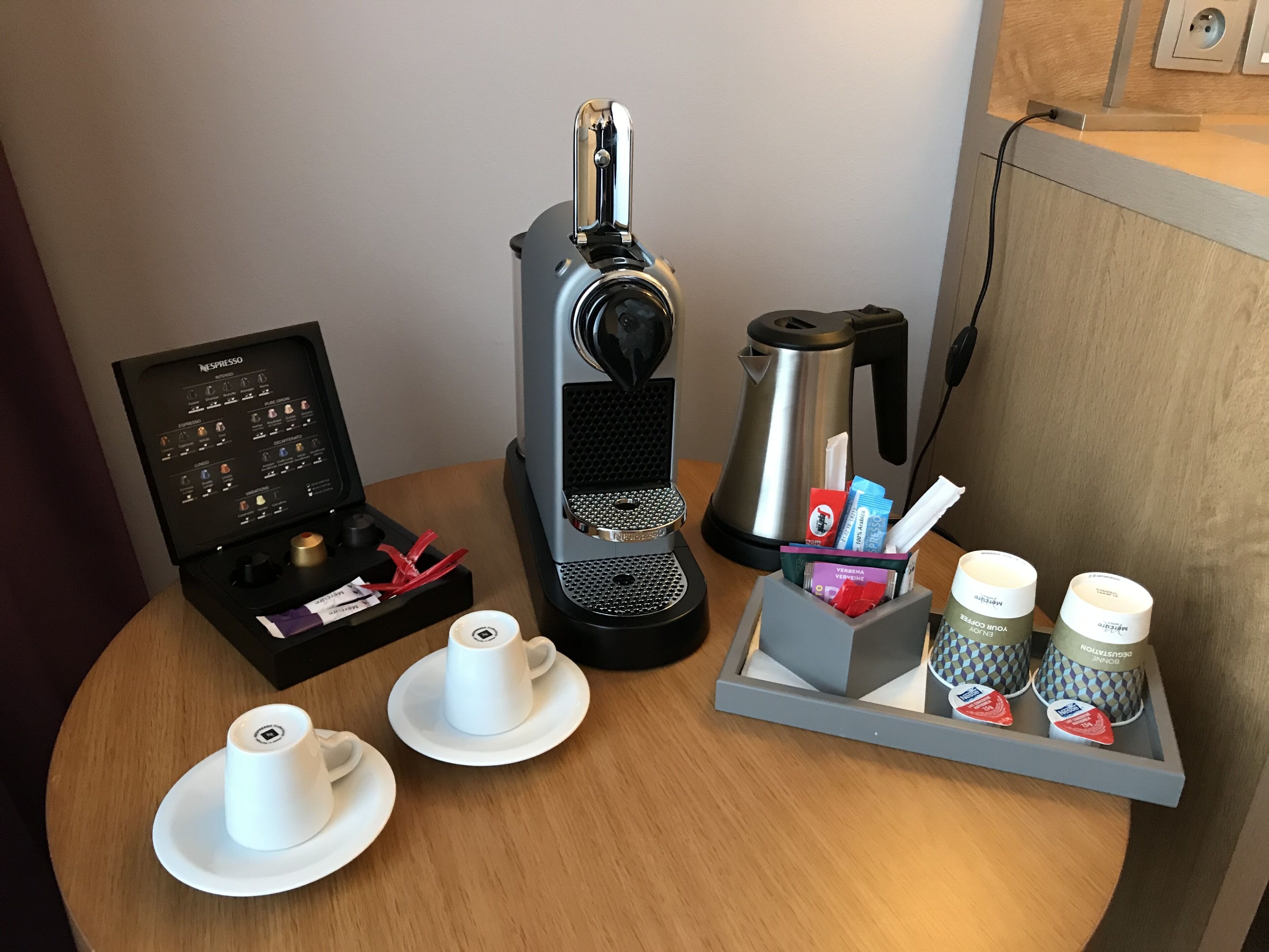 Uma estação de café com máquina Nespresso, cafeteira, xícaras e cápsulas de café sobre uma mesa de madeira.