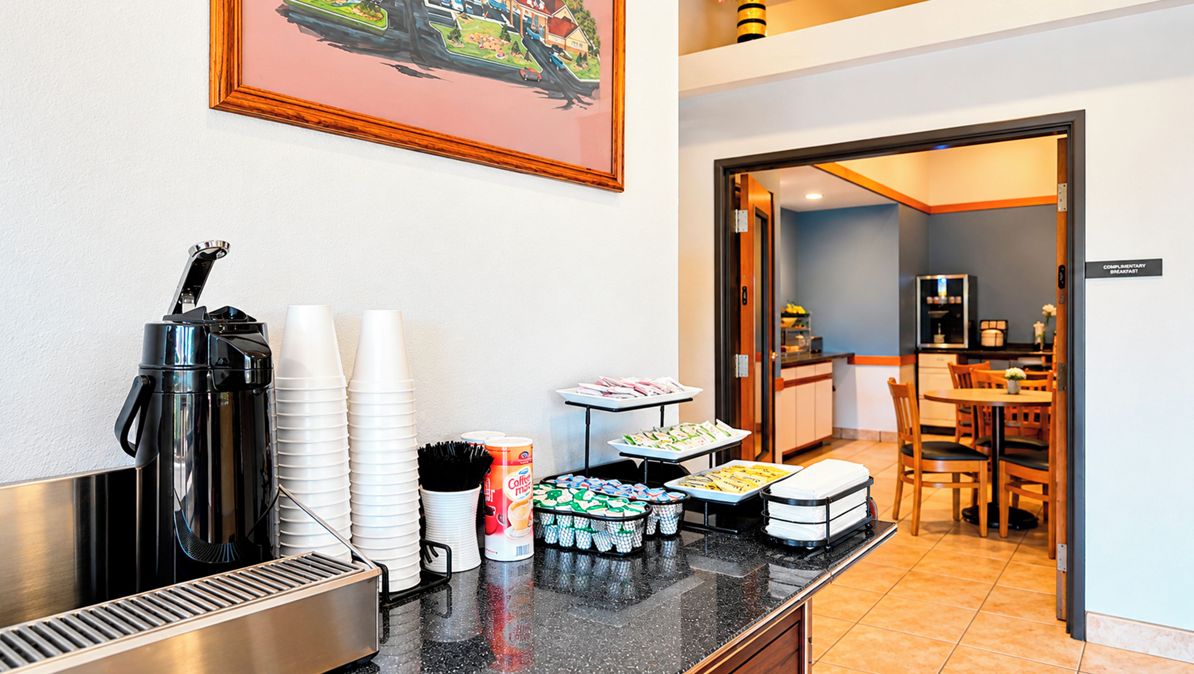 Espaço de café da manhã de hotel, com máquina de café, copos descartáveis e diversas opções para o café da manhã.