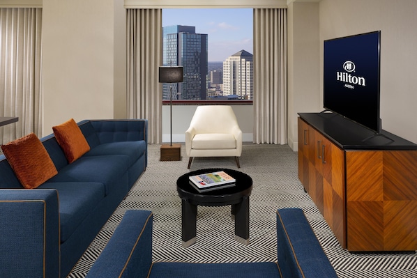 Quarto de hotel moderno com vista para a cidade, um sofá azul, uma poltrona branca, uma mesa de centro preta e um minibar.