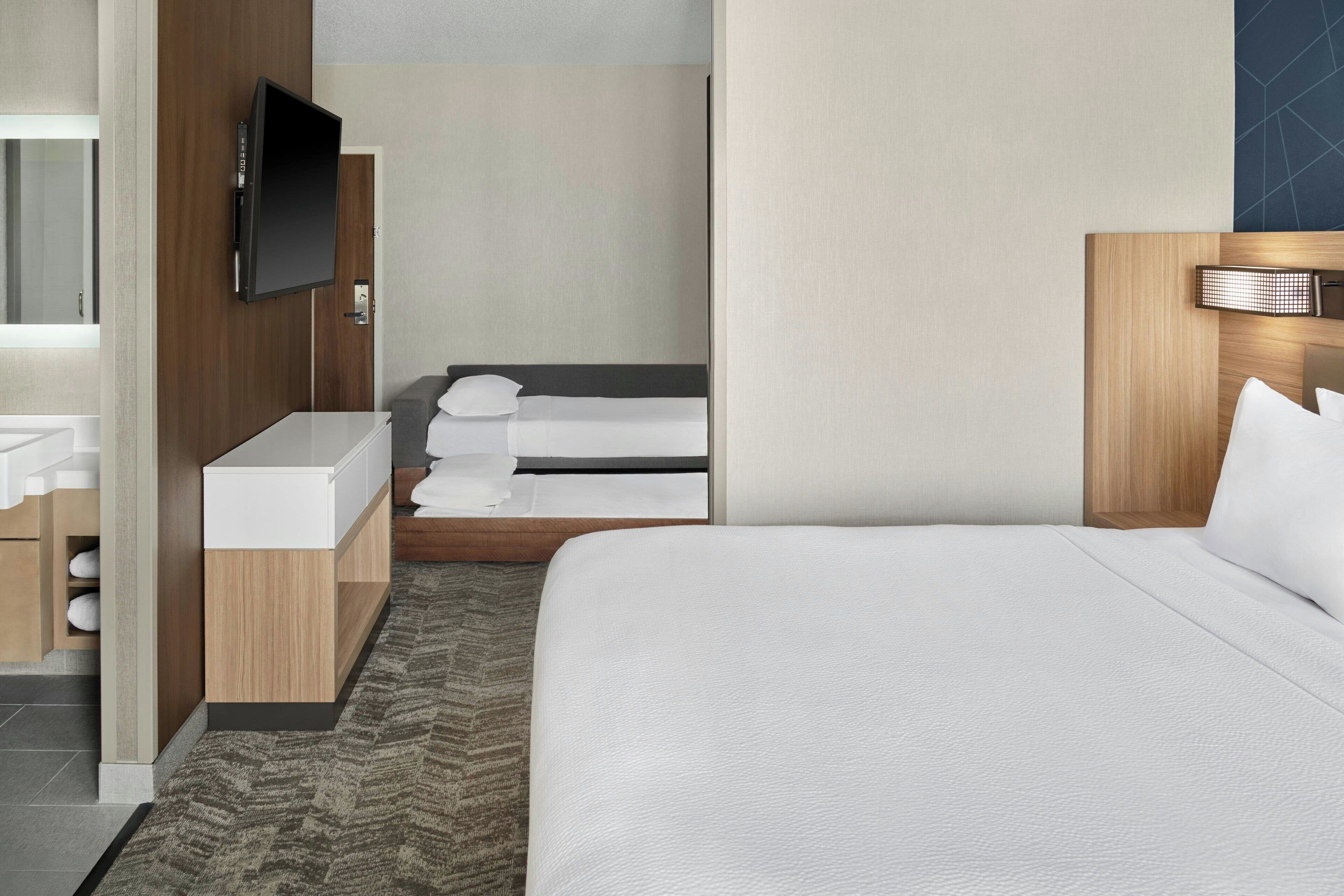 Quarto de hotel com uma cama grande, televisão de tela plana na parede, cabeceira de madeira, um banco embutido com toalha e um espaço pequeno para o banheiro com pia e espelho.
