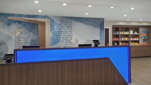 Uma área de recepção de hotel com um balcão de madeira, um painel iluminado em azul e uma parede com diversas palavras e frases.