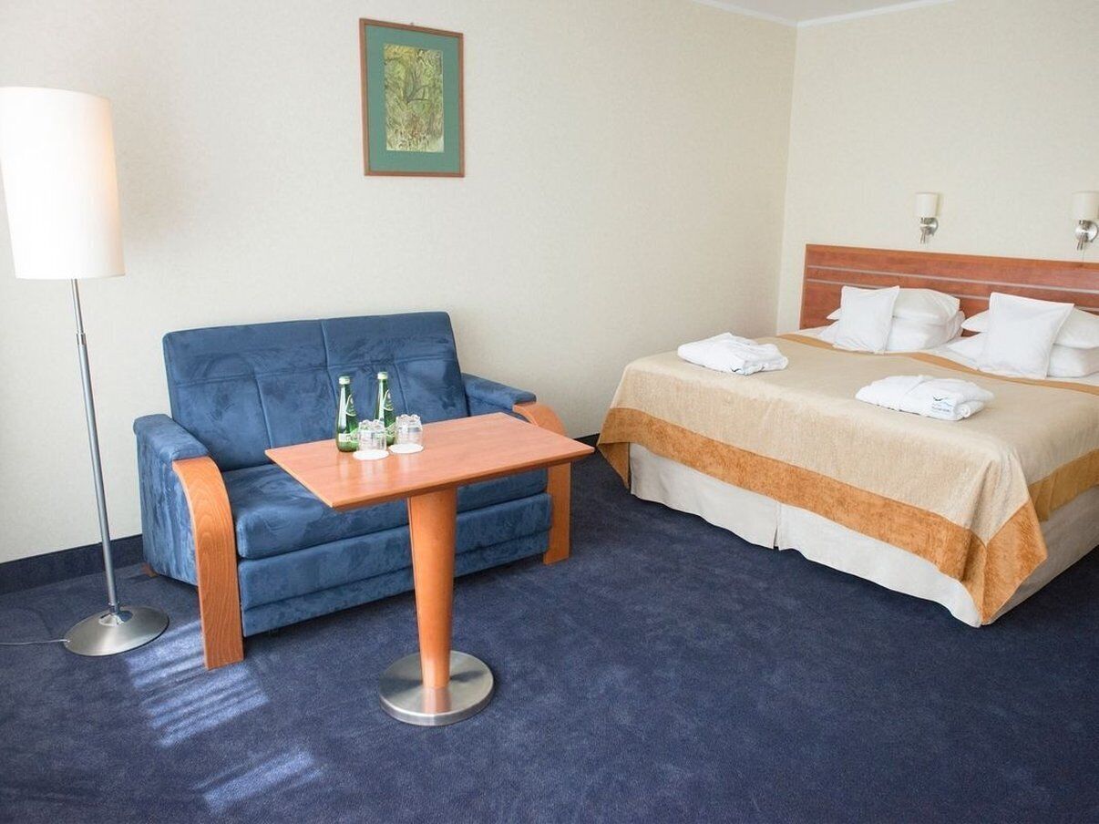 Quarto de hotel com um sofá azul, uma mesa de madeira com uma garrafa e copos, e uma cama com colcha bege e marrom.