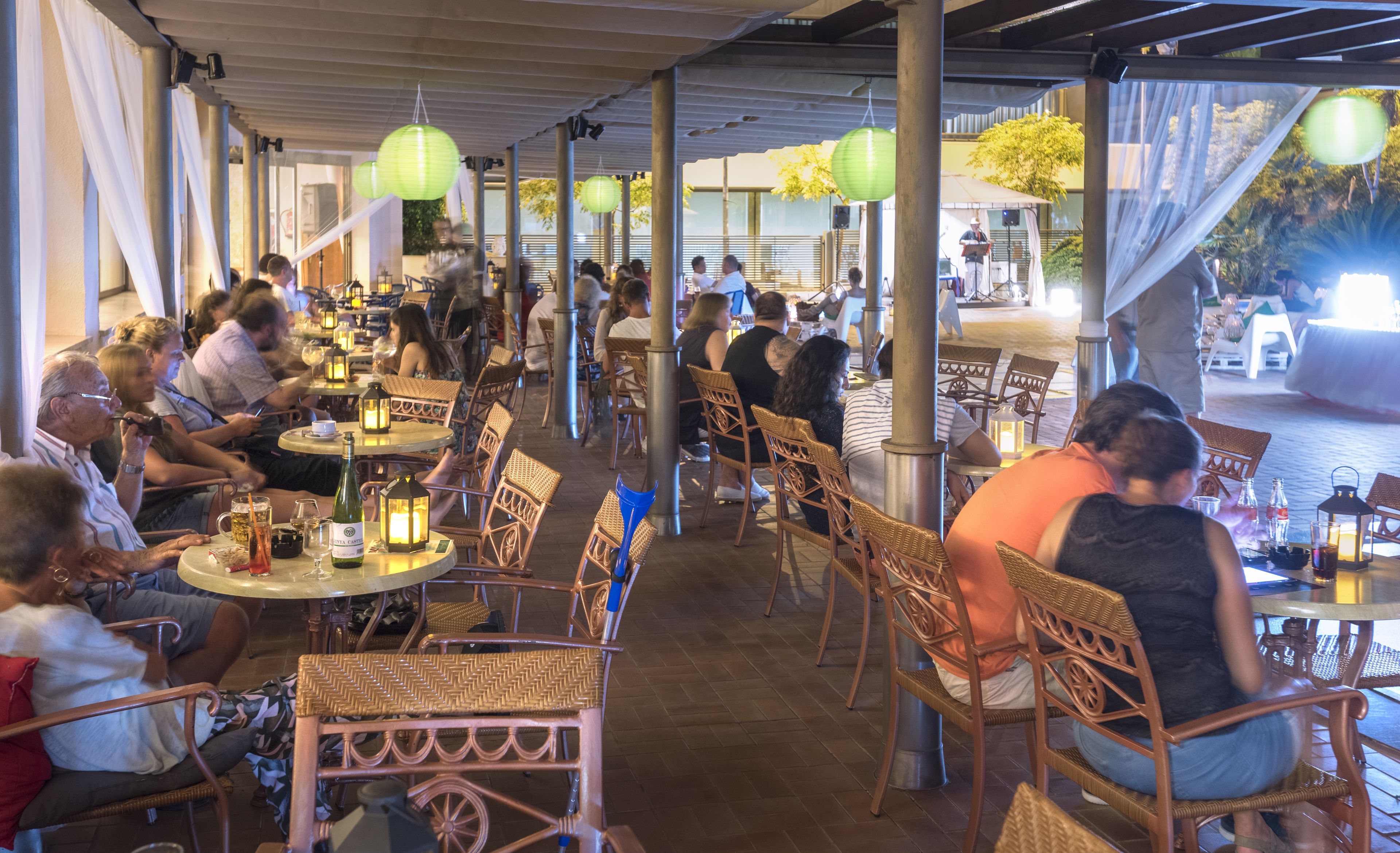 Um café ao ar livre com cadeiras e mesas de vime, lanternas e uma galeria coberta.