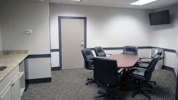 Uma sala de conferências com uma mesa retangular, seis cadeiras de escritório pretas, um quadro branco e uma TV de tela plana fixada na parede.