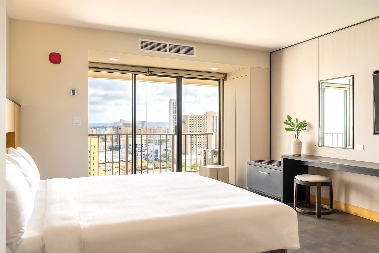 Quarto de hotel com uma cama grande, uma escrivaninha, uma cadeira, uma planta em vaso e vista para a paisagem urbana pelas amplas janelas.