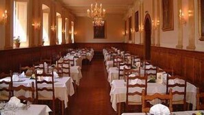 Um salão de jantar comprido e estreito, com fileiras de mesas e cadeiras, um candelabro e quadros nas paredes.
