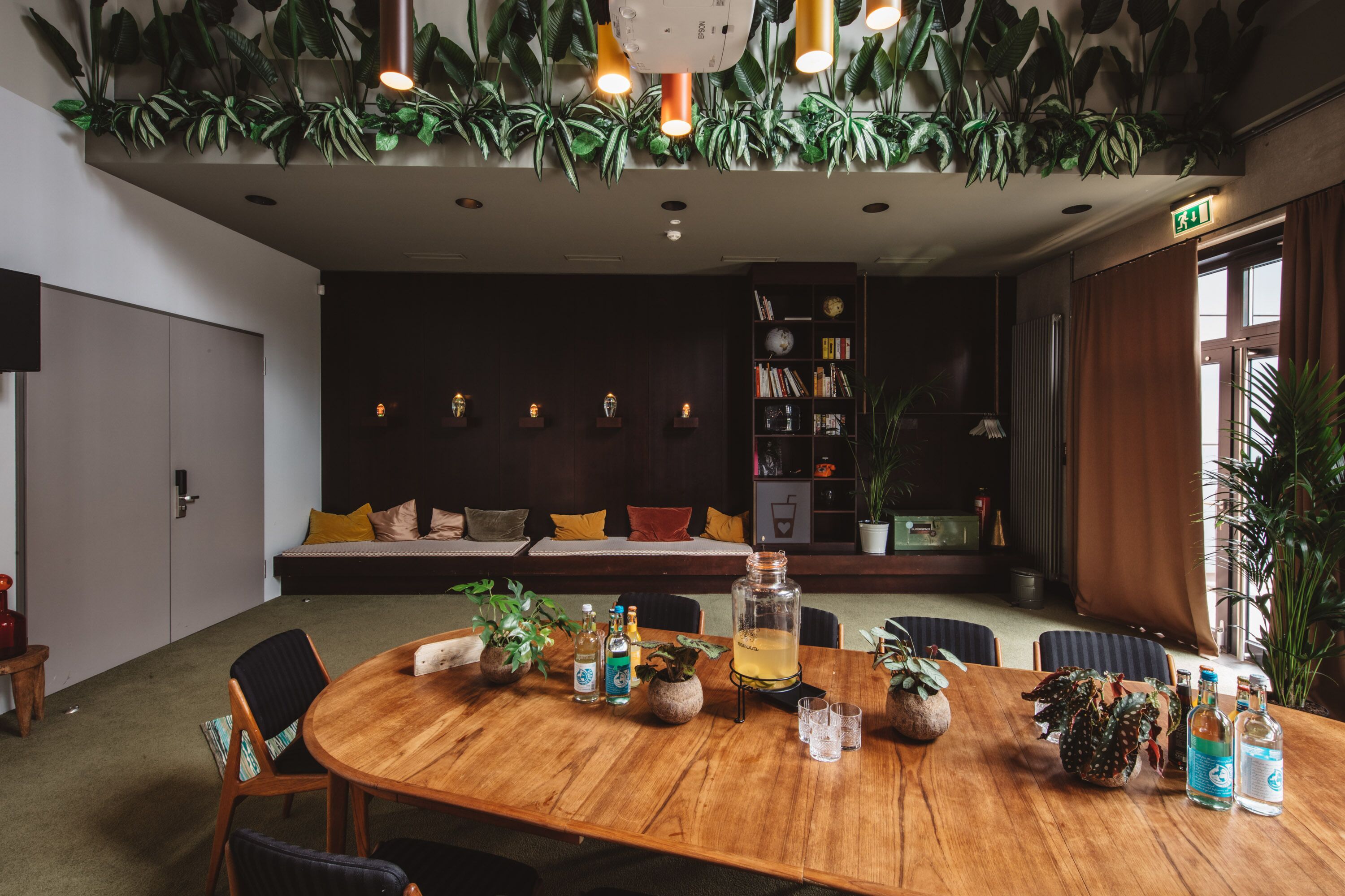 Espaço lounge moderno com mesa de madeira, assentos e teto decorado com plantas verdes.