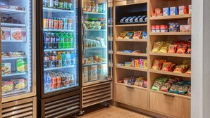 Expositor refrigerado com diversas bebidas e lanches, prateleiras de madeira com produtos embalados e piso cerâmico limpo.