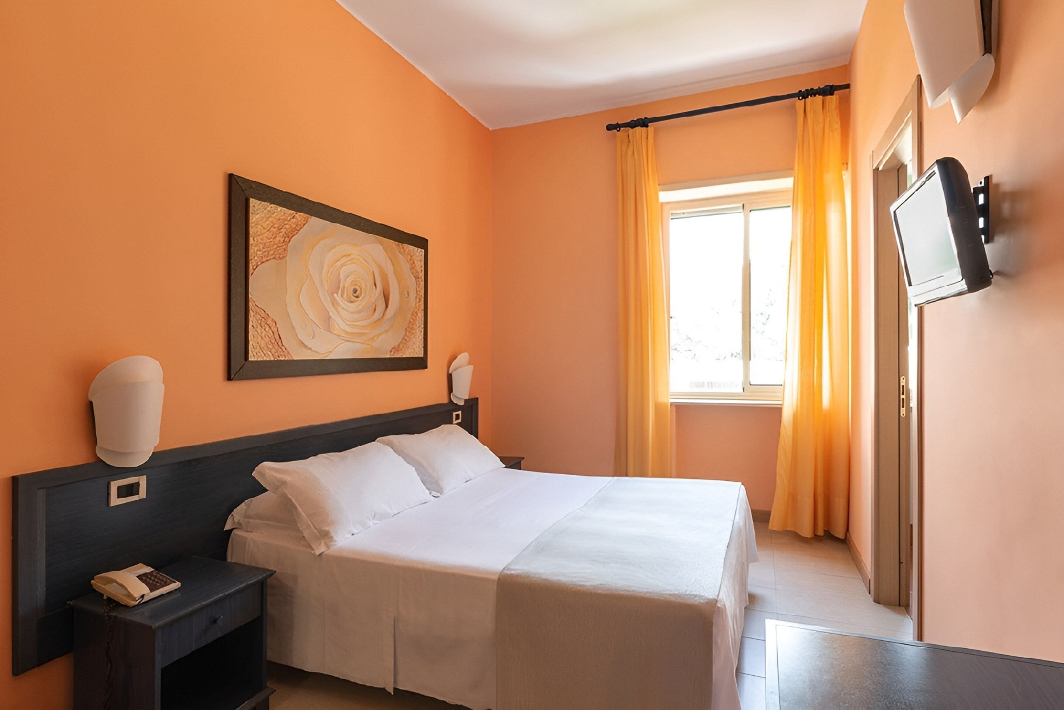 Quarto de hotel com uma cama, uma mesa de cabeceira com telefone, TV fixada na parede e uma janela com cortinas.