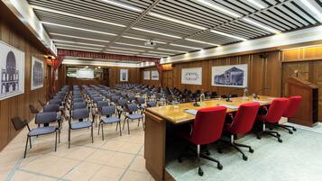 Uma sala de conferências com fileiras de cadeiras, uma mesa comprida com microfones e uma tela de projeção.