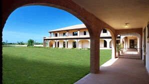 Uma área externa coberta com um arco de tijolos, um gramado e um edifício com várias janelas e portas ao fundo.