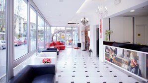 Um saguão moderno de hotel com grandes janelas, balcão de recepção, área de estar e um lustre.