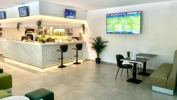Uma área de bar moderna com bancada de mármore, dois bancos, uma televisão exibindo um jogo esportivo e um espaço de estar com uma mesa e cadeiras.
