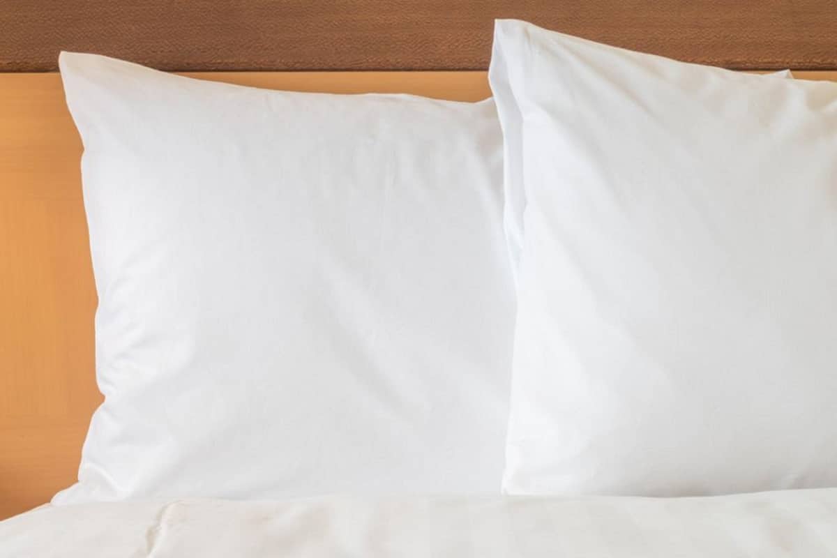 Cama com roupa de cama e almofadas brancas apoiadas em cabeceira de madeira.
