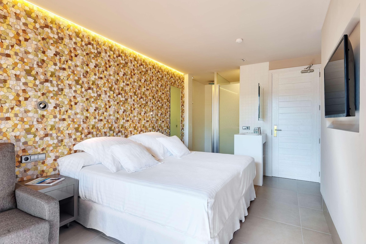 Quarto de hotel moderno com parede em mosaico, uma cama grande com roupa de cama branca, uma mesinha de cabeceira com uma revista e uma TV de tela plana na parede.