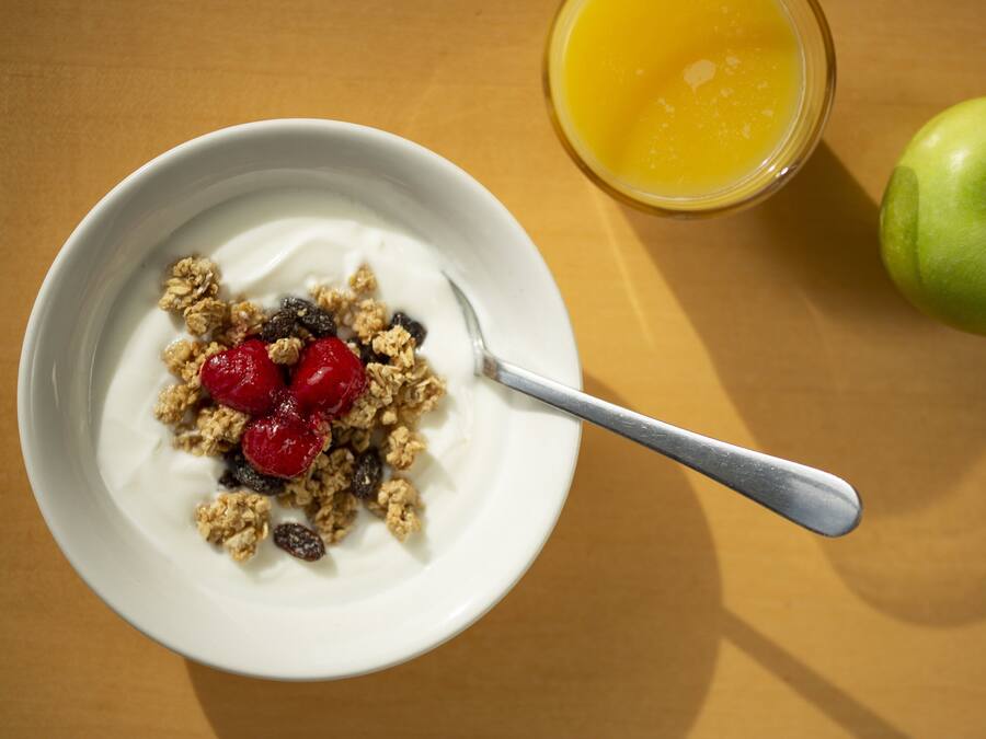 Uma tigela de cereais com leite e fruta, um copo de suco de laranja e uma maçã sobre uma mesa.