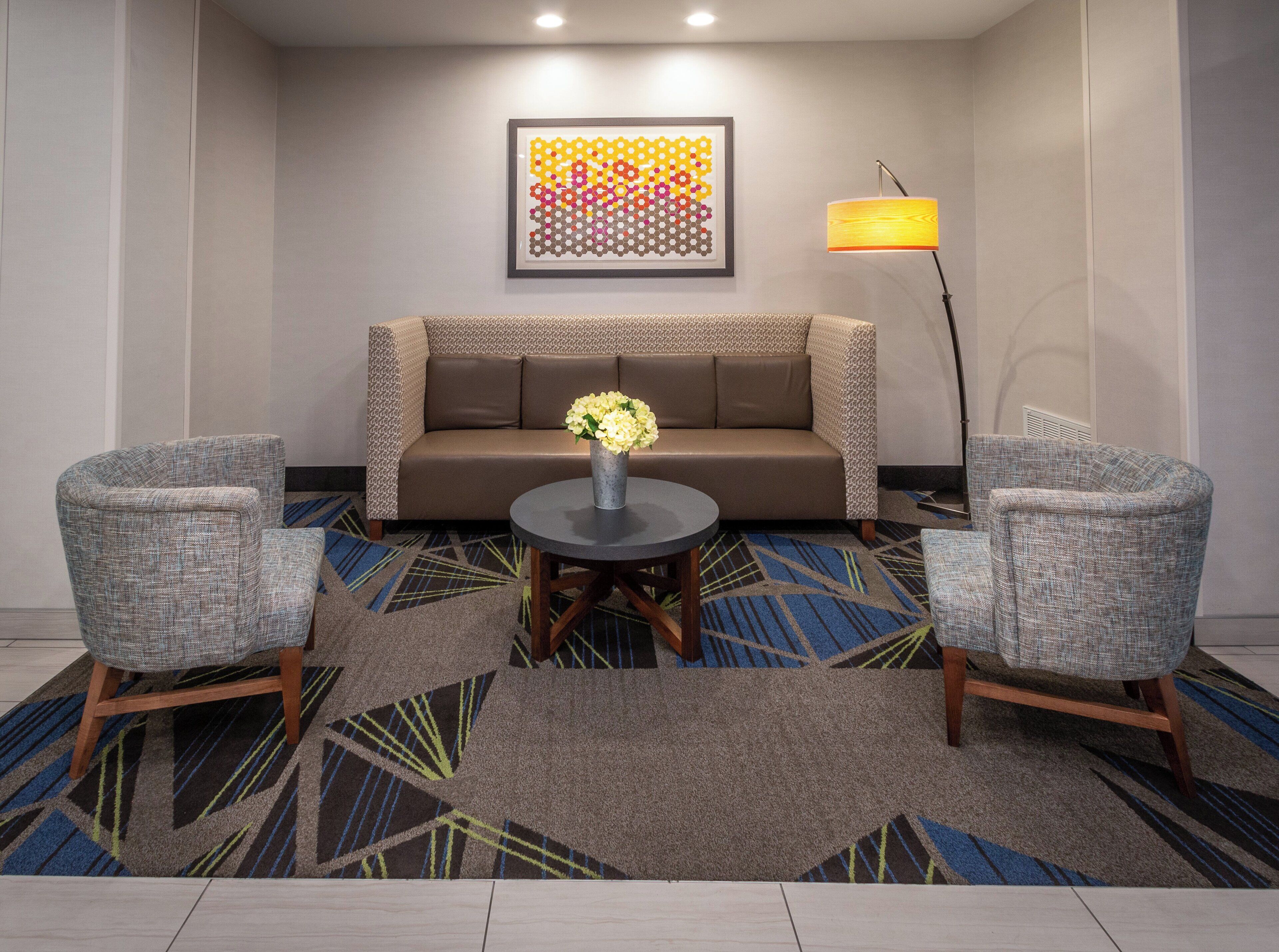 Quarto de hotel com sofá, duas poltronas, mesa redonda pequena com um vaso de flores, luminária de chão e pintura abstrata colorida na parede.