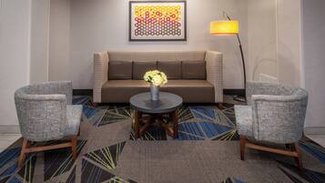 Quarto de hotel com sofá, duas poltronas, mesa redonda pequena com um vaso de flores, luminária de chão e pintura abstrata colorida na parede.