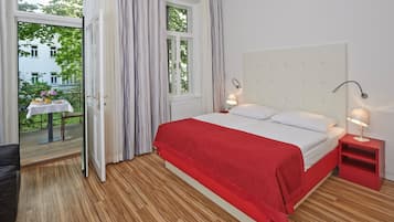 Quarto com cama, luminárias de cabeceira, mesa de cabeceira vermelha, piso de madeira e vista para o verde lá fora.
