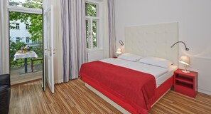 Quarto com cama, luminárias de cabeceira, mesa de cabeceira vermelha, piso de madeira e vista para o verde lá fora.
