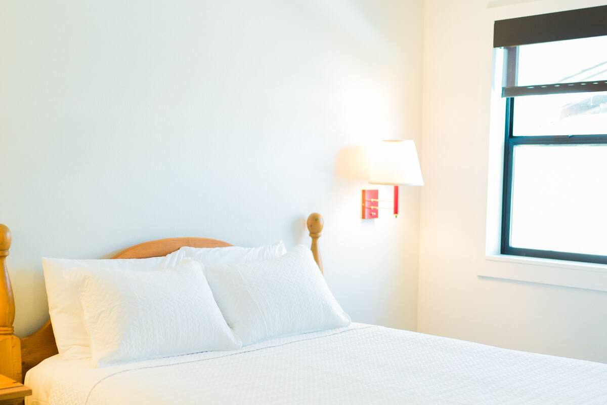 Uma cama bem arrumada com cabeceira de madeira, abajur e janela com persianas.